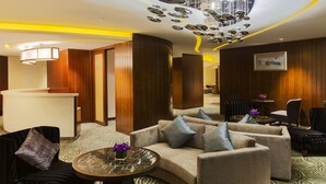 Um saguão de hotel com balcão de recepção, área de estar com sofás e poltronas, um lustre e um tapete estampado.