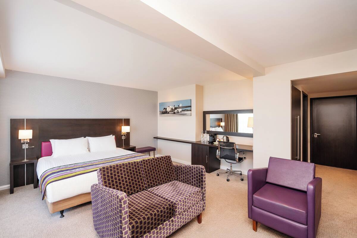 Quarto de hotel com uma cama grande, uma escrivaninha, uma poltrona roxa e um sofá estampado.