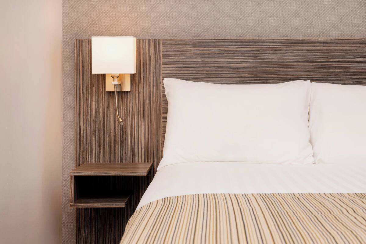 Quarto de hotel com uma cama grande, uma mesa de cabeceira com abajur e cabeceira de madeira.