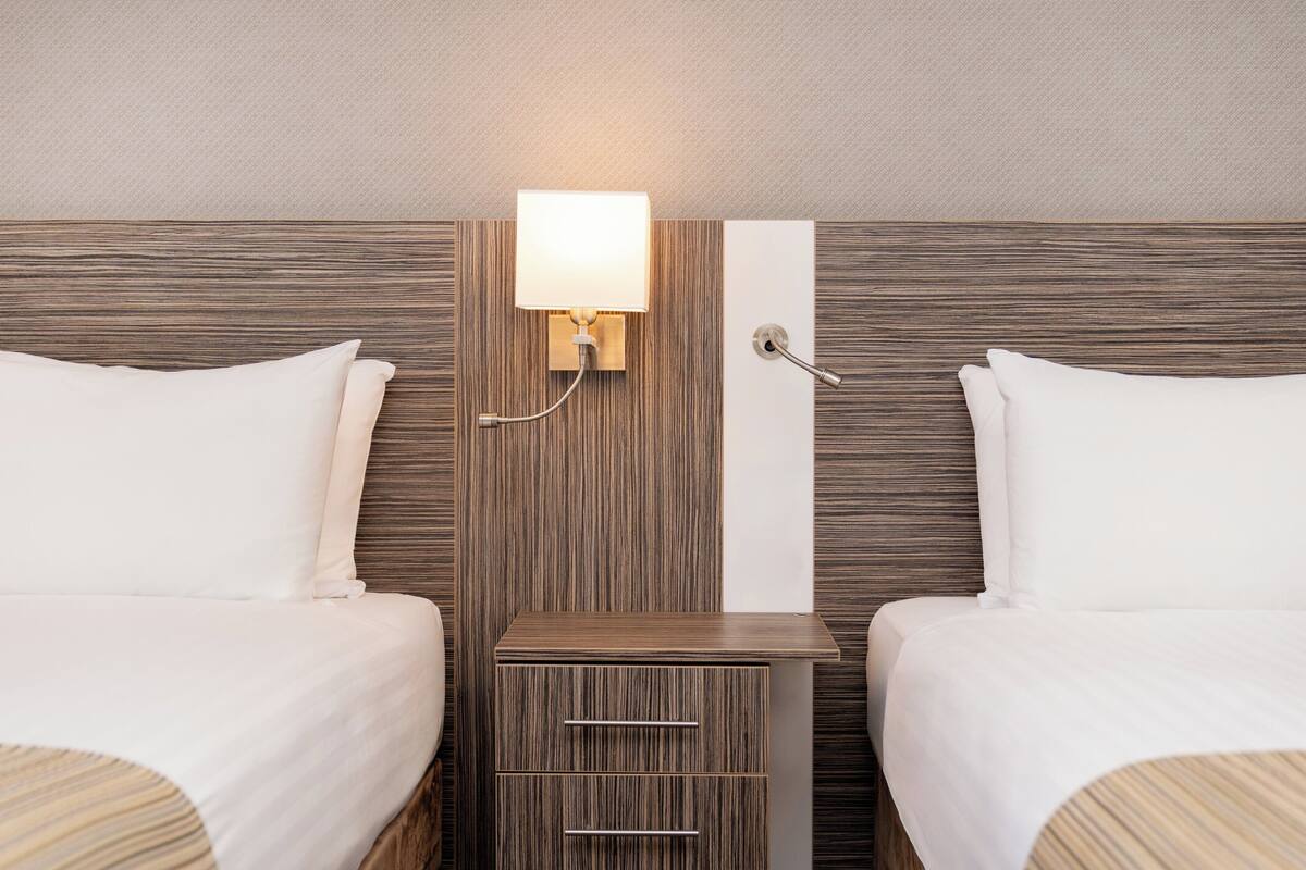 Quarto de hotel com duas camas, uma mesa de cabeceira, uma luminária na parede e um porta-toalhas.