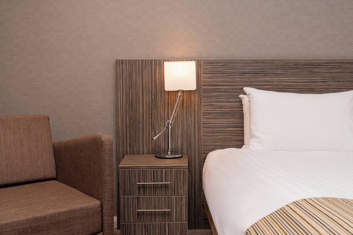 Quarto de hotel com uma cama, mesa de cabeceira com abajur e um espaço para sentar.