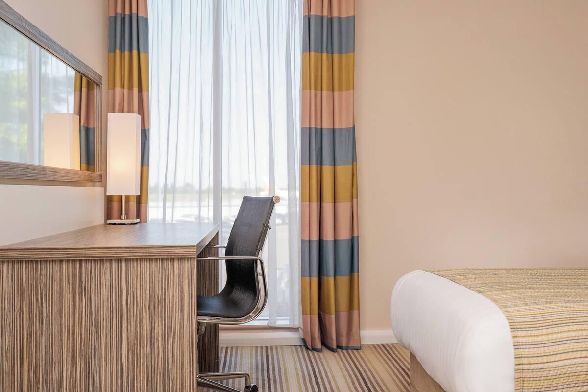 Quarto de hotel com escrivaninha de madeira, uma cadeira, uma cama, janela com cortinas e uma luminária.