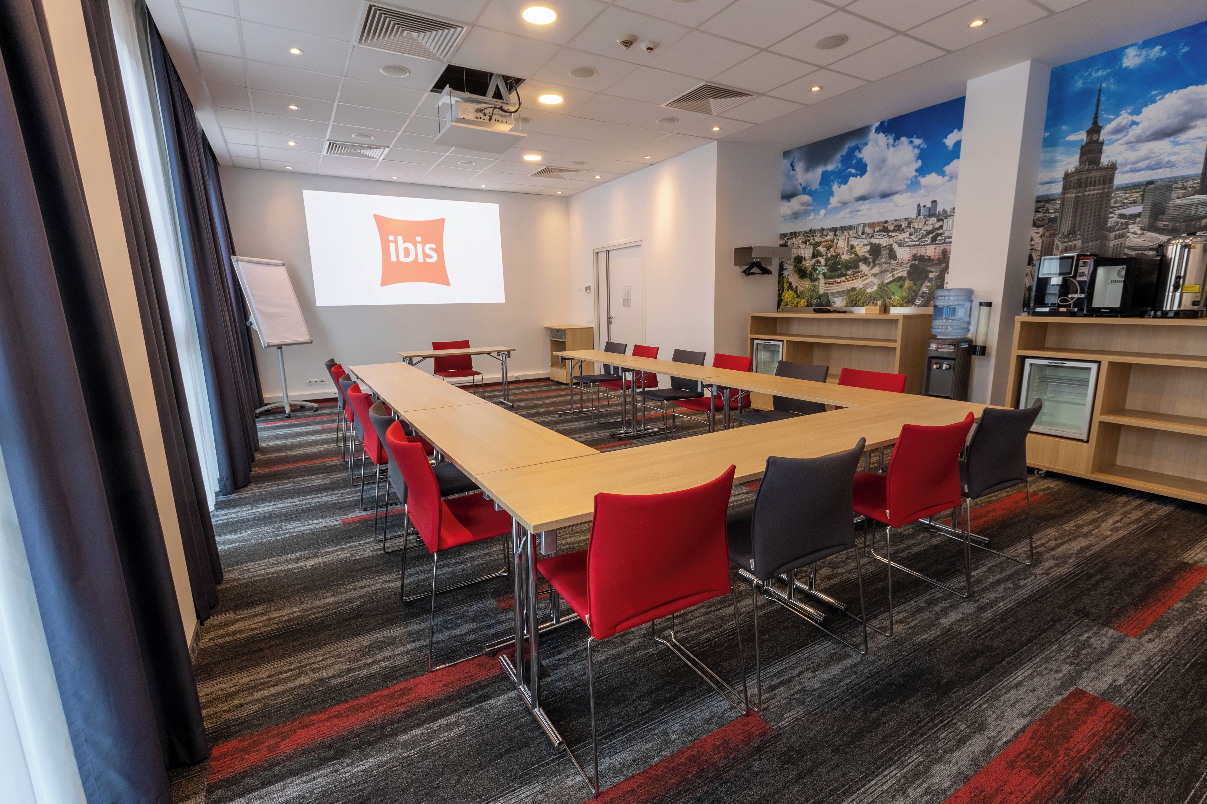 Sala de conferências com uma mesa comprida, cadeiras vermelhas, uma tela de projetor exibindo 'ibis' e um mural com vista para a cidade.