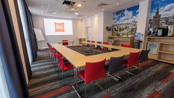 Sala de conferências com uma mesa comprida, cadeiras vermelhas, uma tela de projetor exibindo 'ibis' e um mural com vista para a cidade.