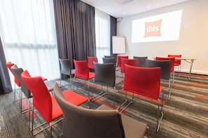 Sala de conferência com tela de projeção exibindo o logotipo 'ibis', disposição de assentos e piso coberto por carpete.