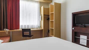 Quarto de hotel com cama, escrivaninha com cadeira, televisão, guarda-roupa e janela com cortinas.