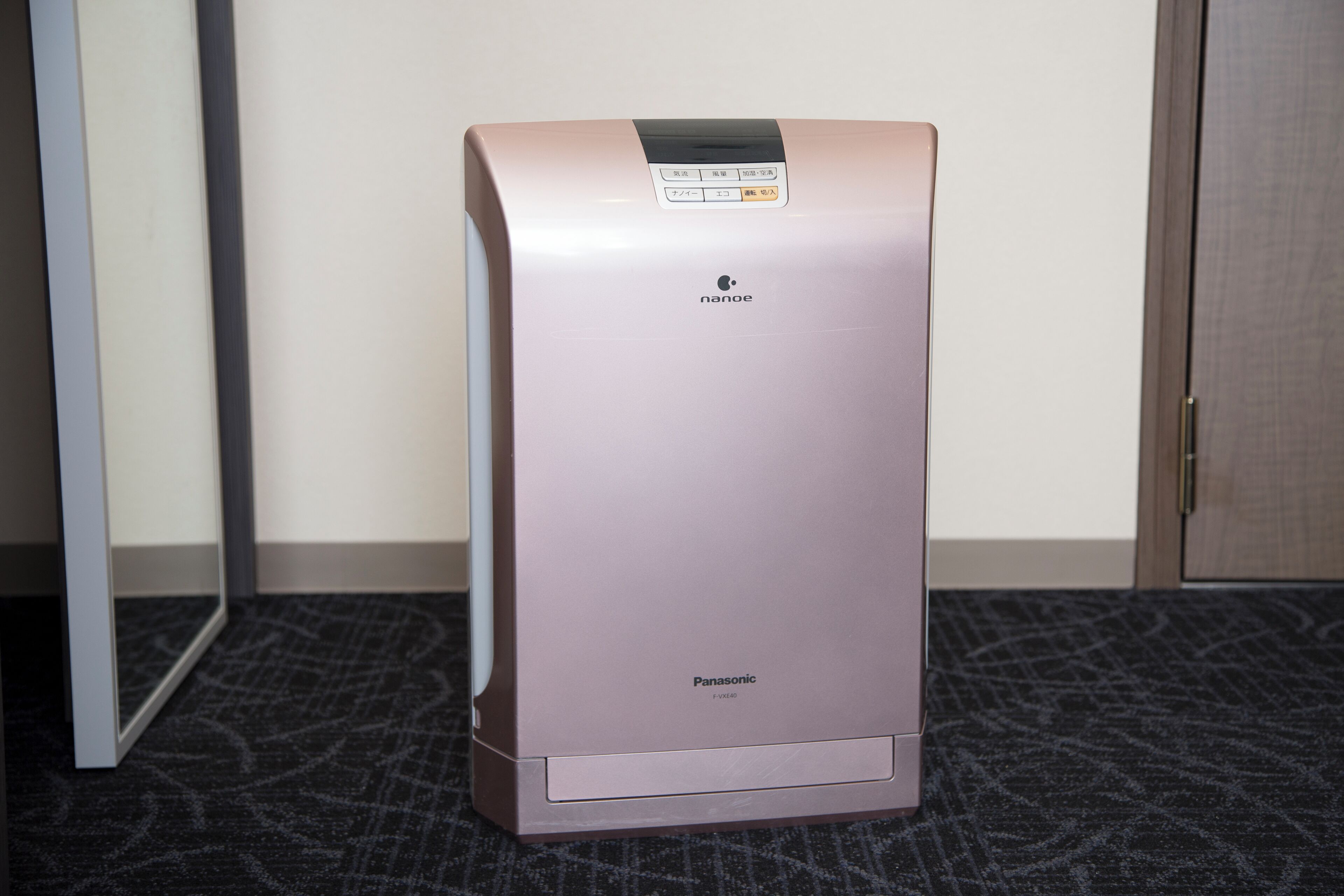 Um purificador de ar Panasonic num quarto com carpete estampado e uma porta ao fundo.