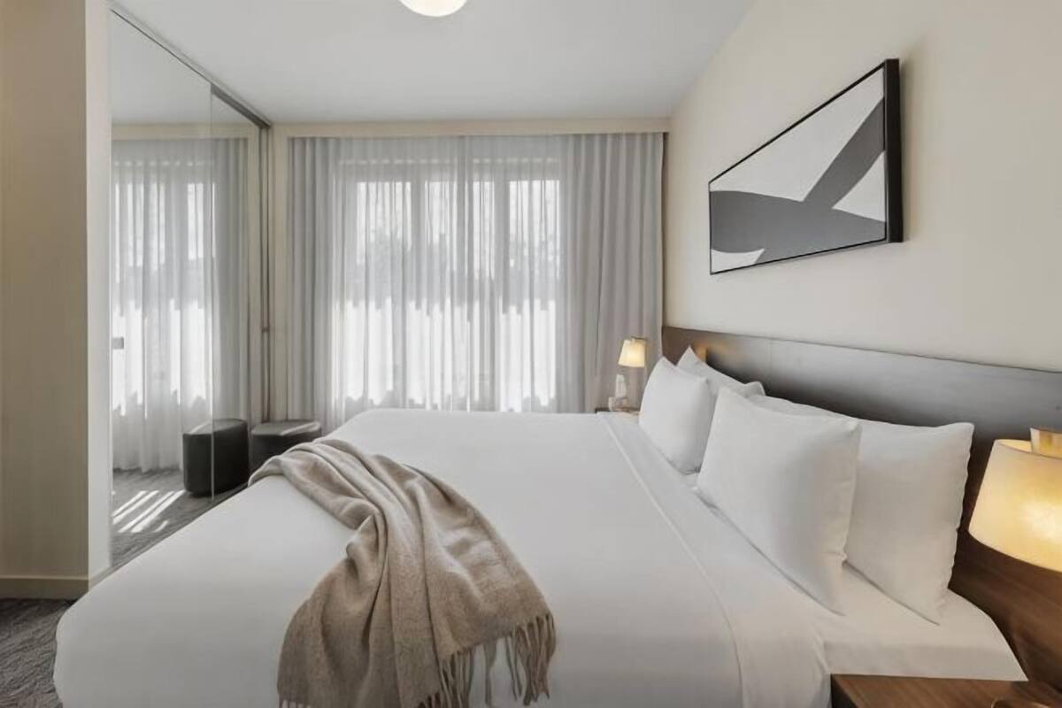 Quarto de hotel com uma cama grande, uma mesa de cabeceira com abajur, vista para uma janela com cortinas translúcidas e um quadro abstrato moderno na parede.
