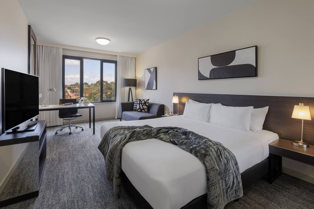 Quarto de hotel com uma cama grande, uma mesa de trabalho com televisão, uma cadeira e vista para a cidade.