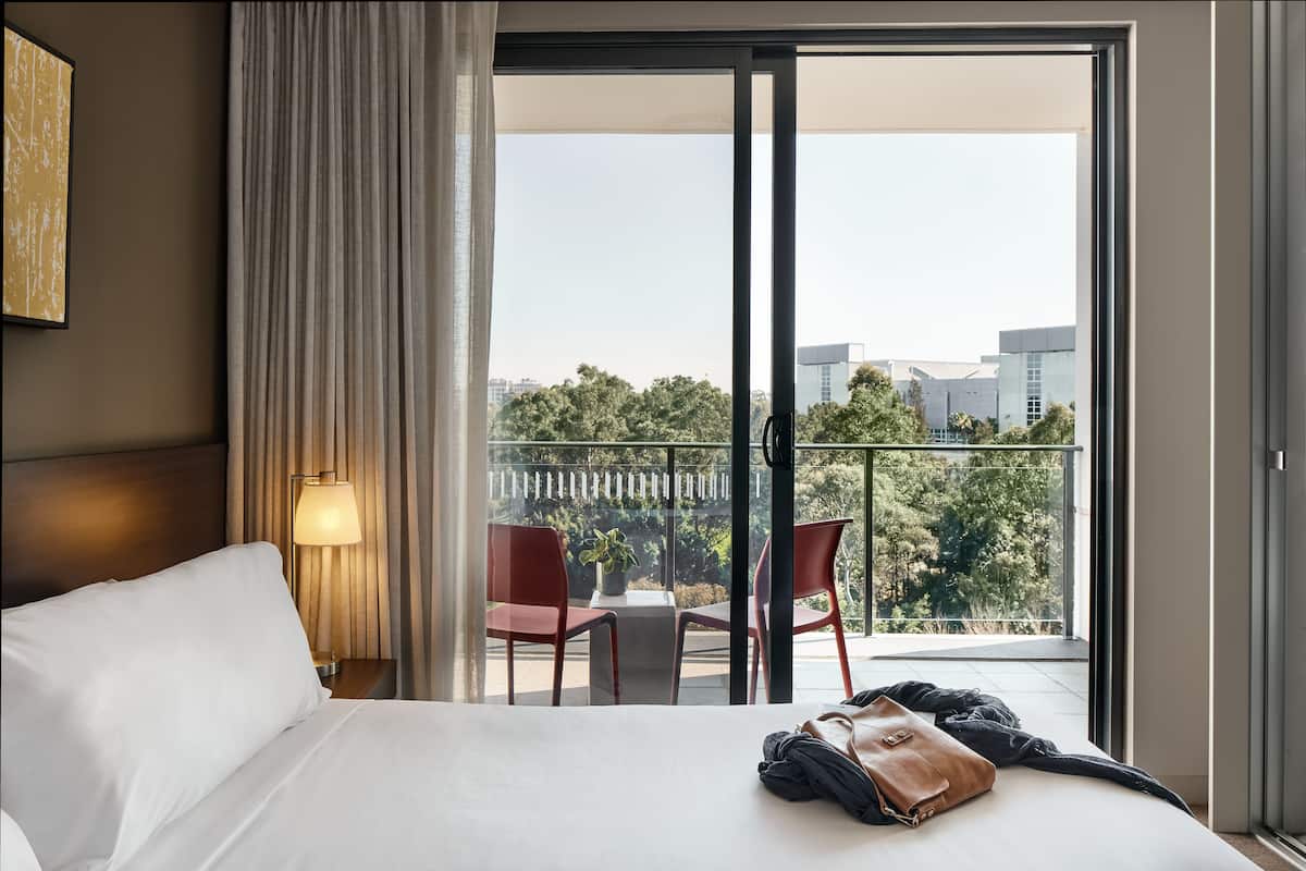 Quarto de hotel com cama, mesa de cabeceira com abajur, mesa com cadeiras e varanda com vista para árvores e edifícios.