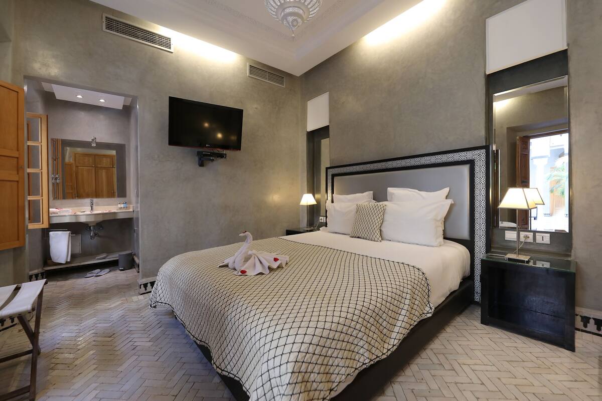Quarto de hotel com uma cama grande, uma TV de tela plana na parede, um banheiro com pia e espelho, e um banco de madeira.