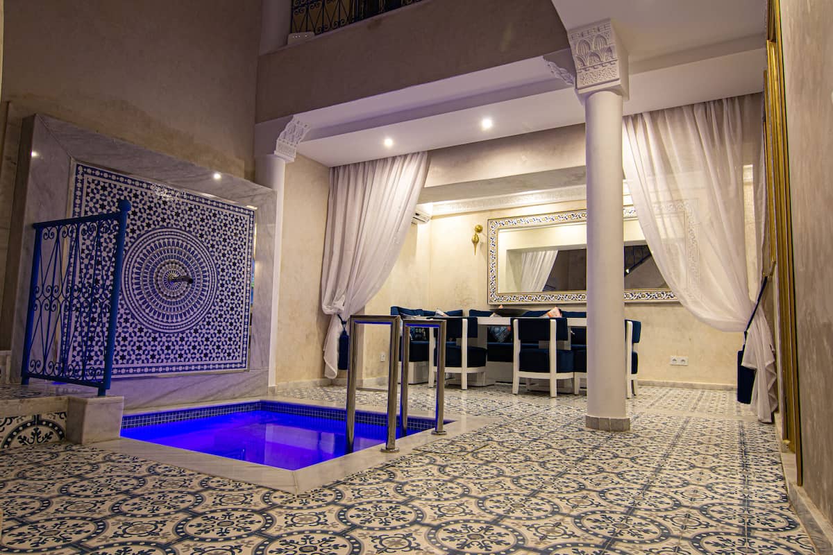 Uma área luxuosa com piscina interna, revestida de azulejos azuis, detalhes ornamentais em azulejo e um espaço de estar com cadeiras azuis e cortinas brancas.