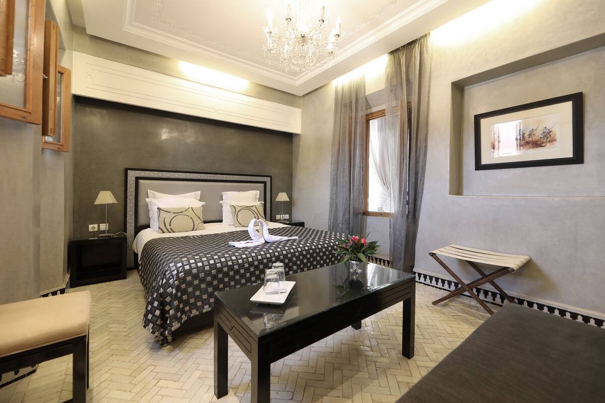 Quarto de hotel com uma cama grande, uma mesinha de cabeceira, uma cadeira, uma pequena mesa, um candelabro e um quadro na parede.