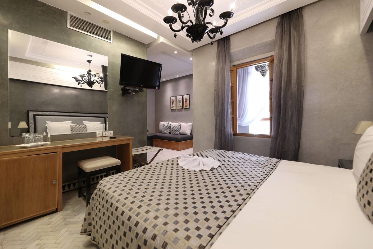 Quarto de hotel moderno com uma cama grande, uma mesa com cadeira, televisão na parede e janela com cortinas.