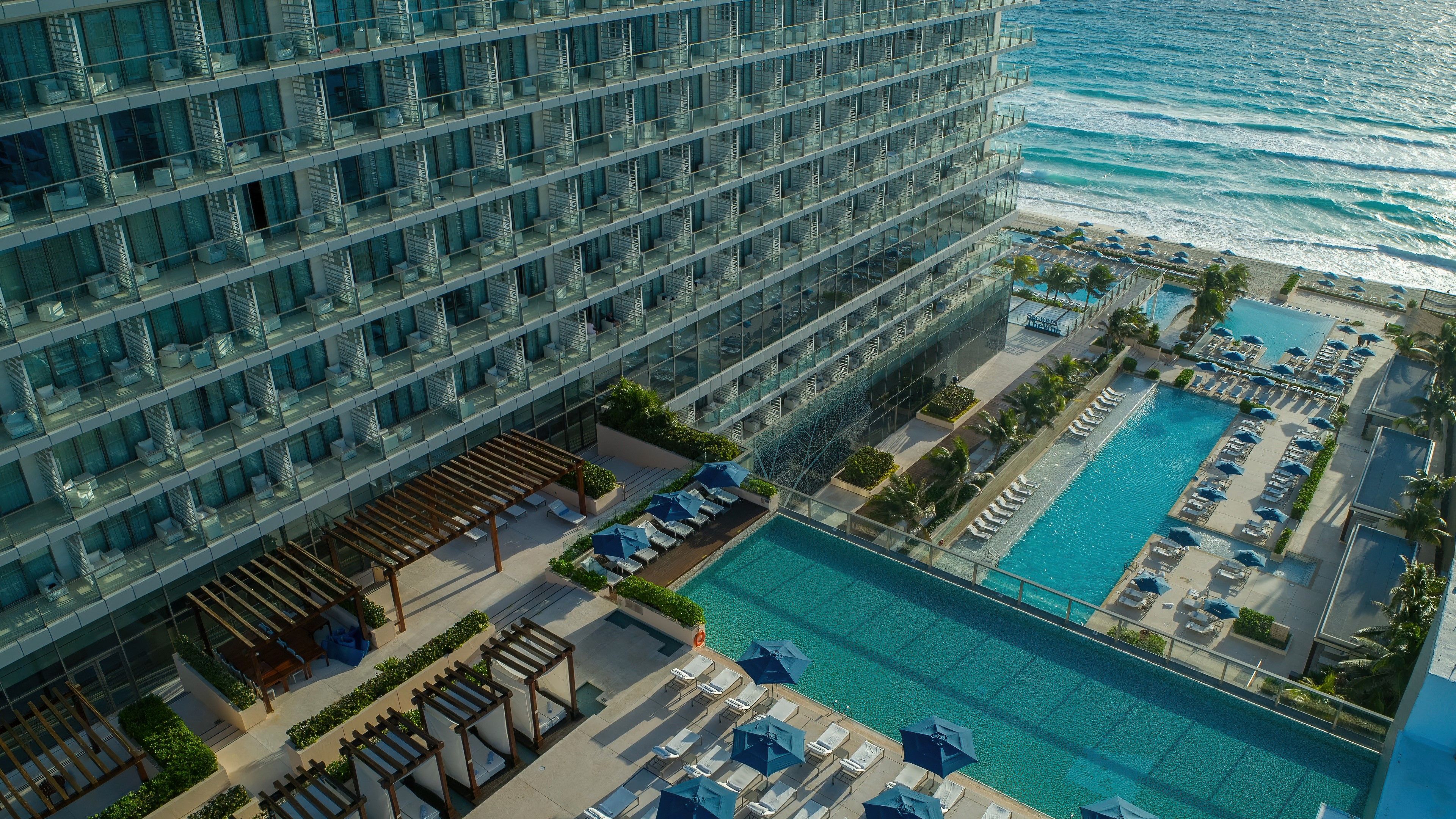 Hotel alto com várias varandas, área de piscina com espreguiçadeiras e guarda-sóis, e vista para o oceano.