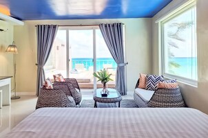 Quarto de hotel com uma cama, duas cadeiras de vime, uma mesinha com uma planta e vista para o mar através de uma porta de correr.