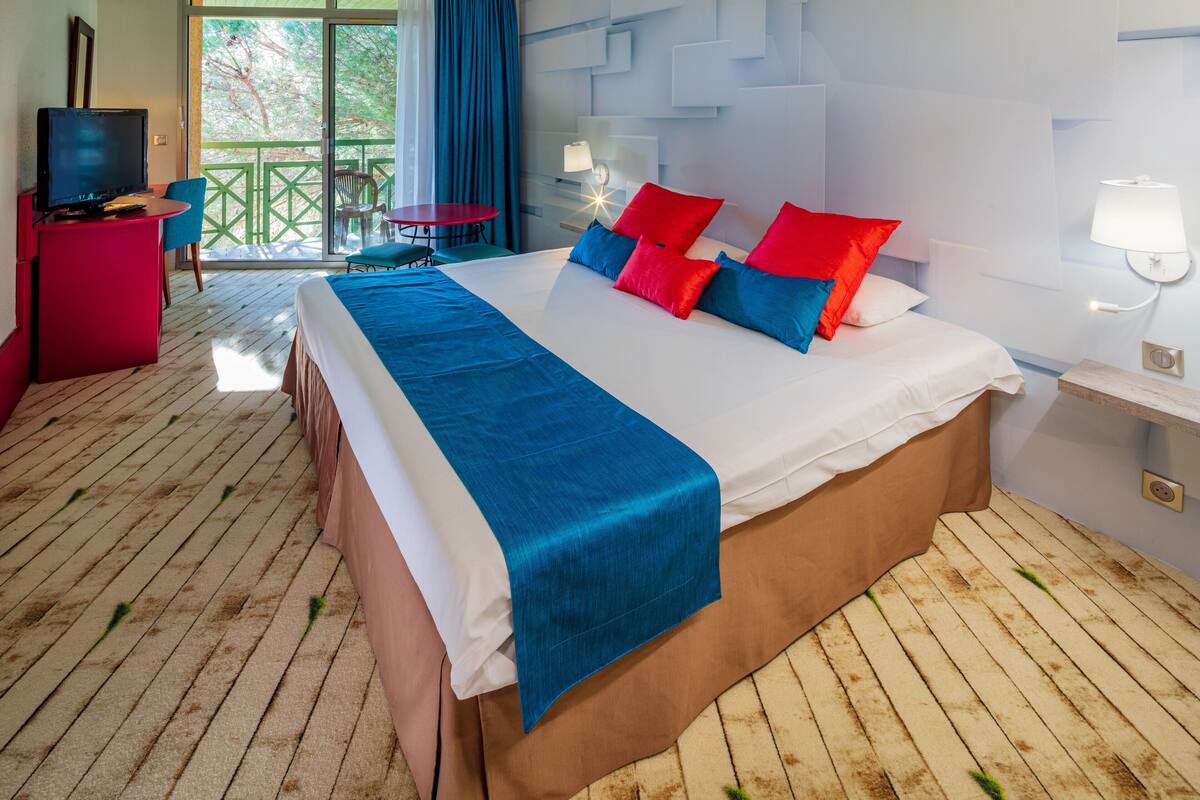 Quarto de hotel com uma cama grande, uma escrivaninha com televisão, uma varanda com mesa e cadeiras, e vista para árvores.