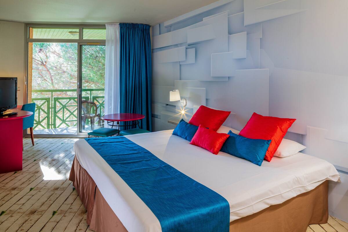 Quarto de hotel com uma cama grande, uma escrivaninha, uma cadeira e uma varanda com mesa e cadeiras.