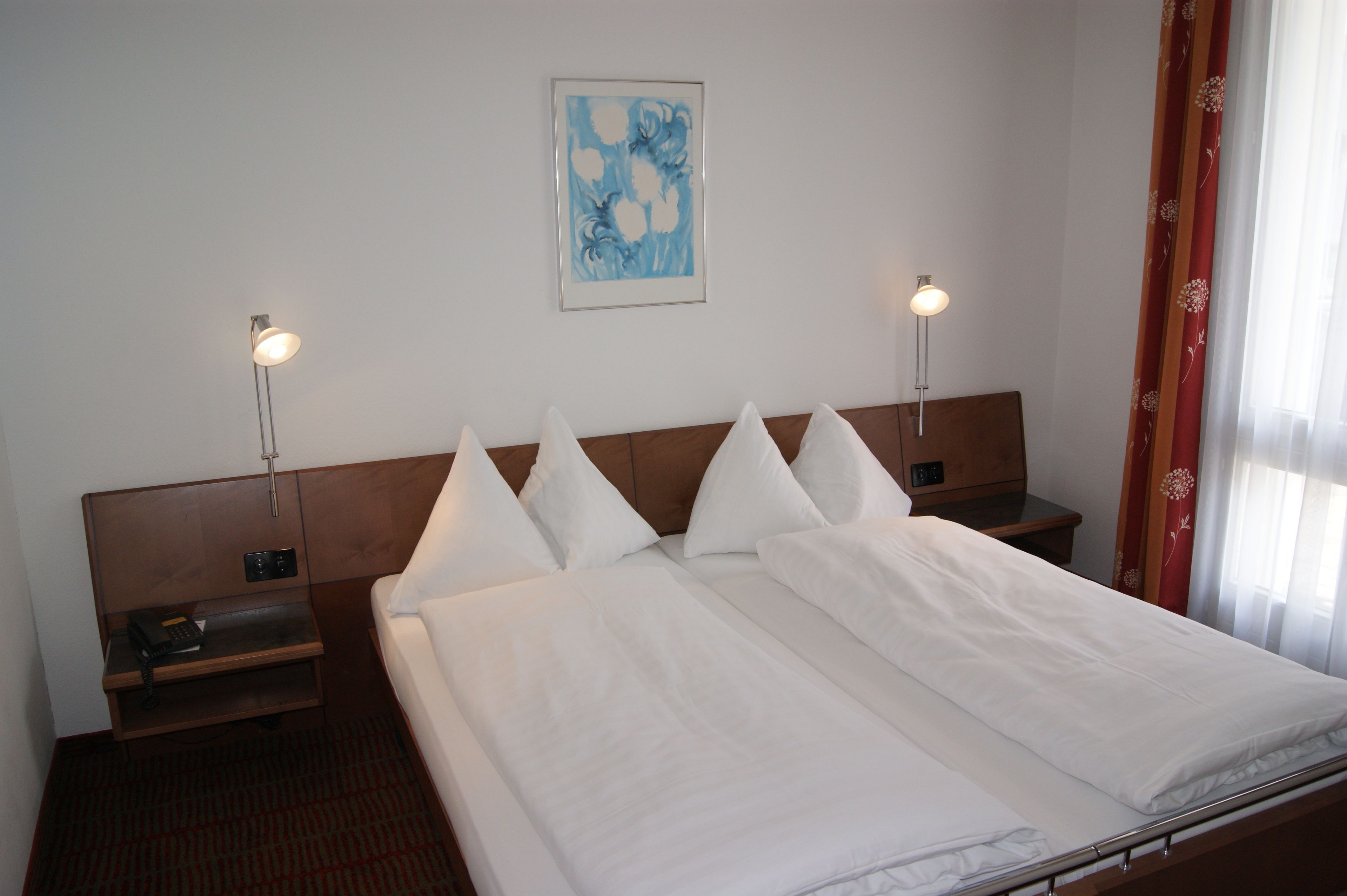 Quarto de hotel com uma cama grande, duas mesinhas de cabeceira, um criado-mudo, um quadro pendurado na parede e uma janela com cortinas.