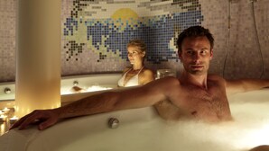 Um homem e uma mulher numa banheira, com um painel de mosaicos ao fundo.