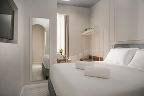 Quarto de hotel com uma cama grande, uma mesa de cabeceira, um espelho, um cabideiro e uma televisão fixada na parede.