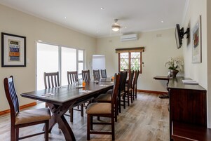 Uma sala de jantar com uma mesa de madeira comprida, várias cadeiras e uma pequena mesa lateral com um vaso de flores.