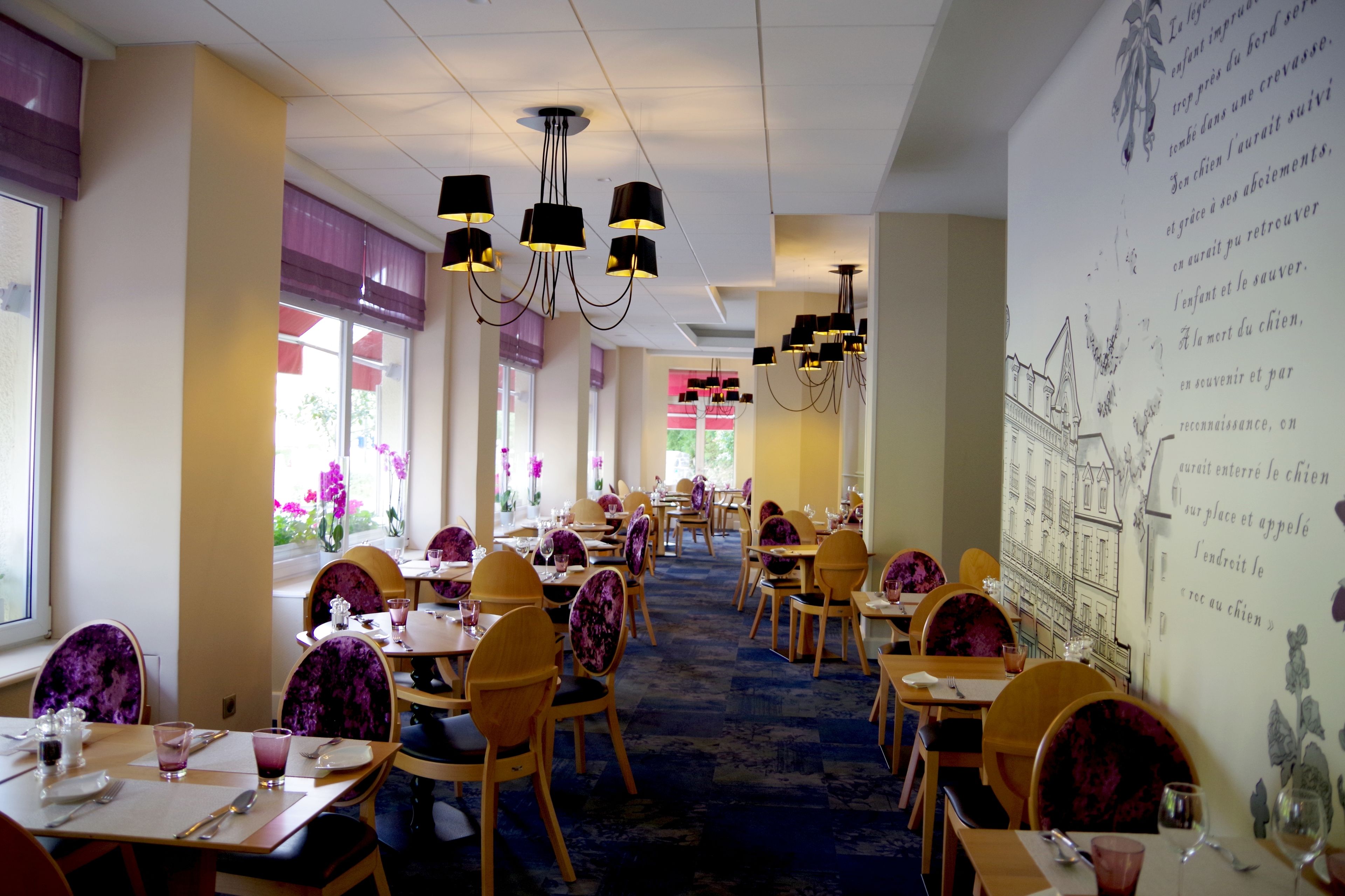 Um restaurante com várias mesas e cadeiras, janelas amplas e luminárias pendentes.