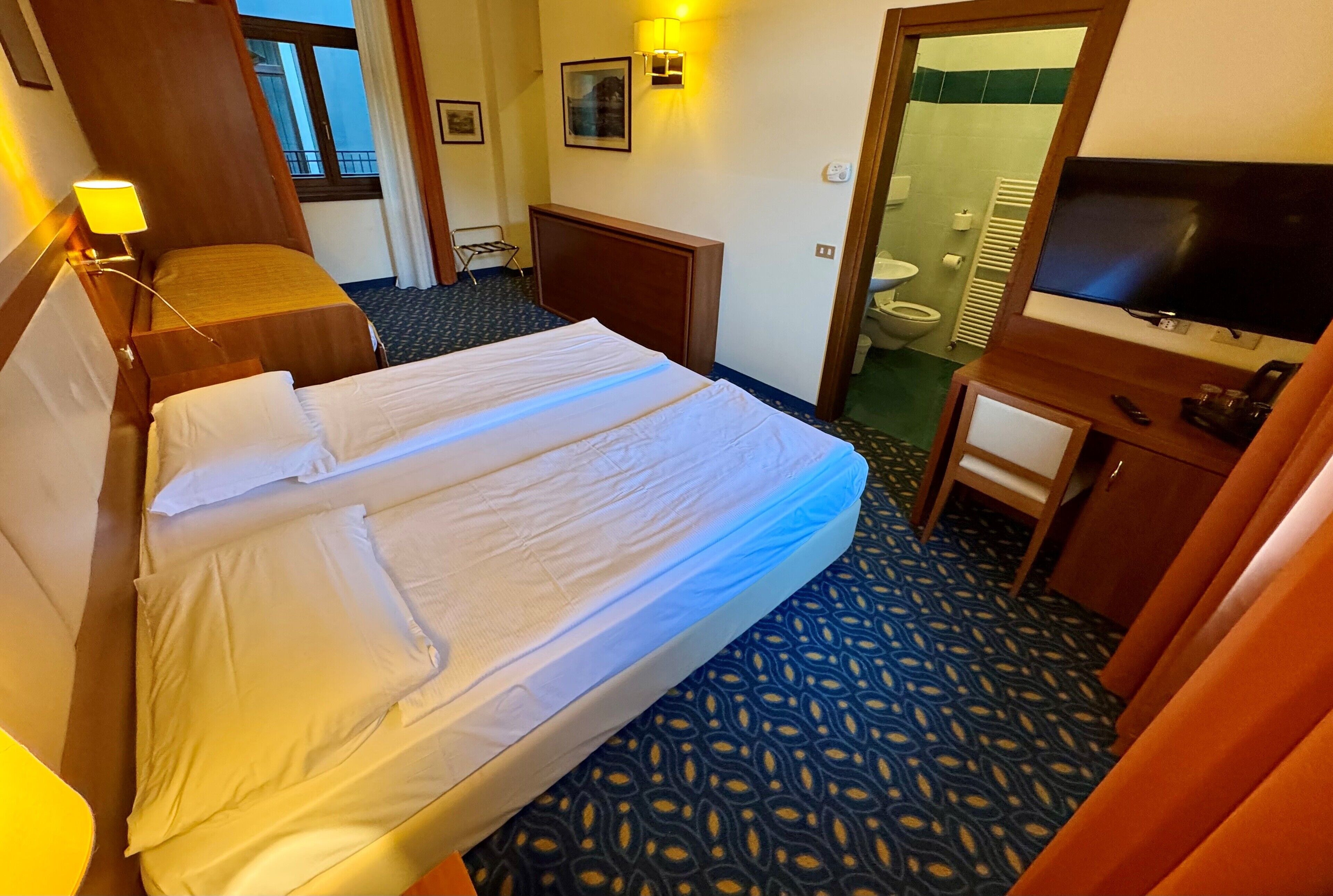 Quarto de hotel com cama de casal, uma escrivaninha com cadeira, televis ão e banheiro visível por uma porta aberta.