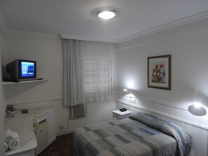 Quarto com cama, televisão em uma estante, mesa de cabeceira com abajur e quadro na parede.