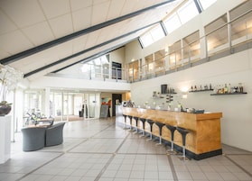 Um hall amplo com teto alto, área de bar central com banquetas e escada que leva a um andar superior.