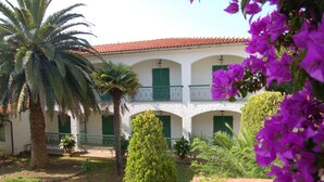 Um edifício com fachada branca, caixilhos verdes e telhado de telhas vermelhas. Possui várias varandas com grades verdes, e o edifício é rodeado por vegetação, incluindo palmeiras e plantas com flores.