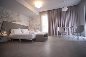 Quarto de hotel moderno com cama, mesa de cabeceira, abajur e uma área de refeições com cadeiras e mesa.
