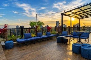 Terraço no telhado com piso de madeira, assentos acolchoados azuis, uma mesa redonda e plantas em vasos.
