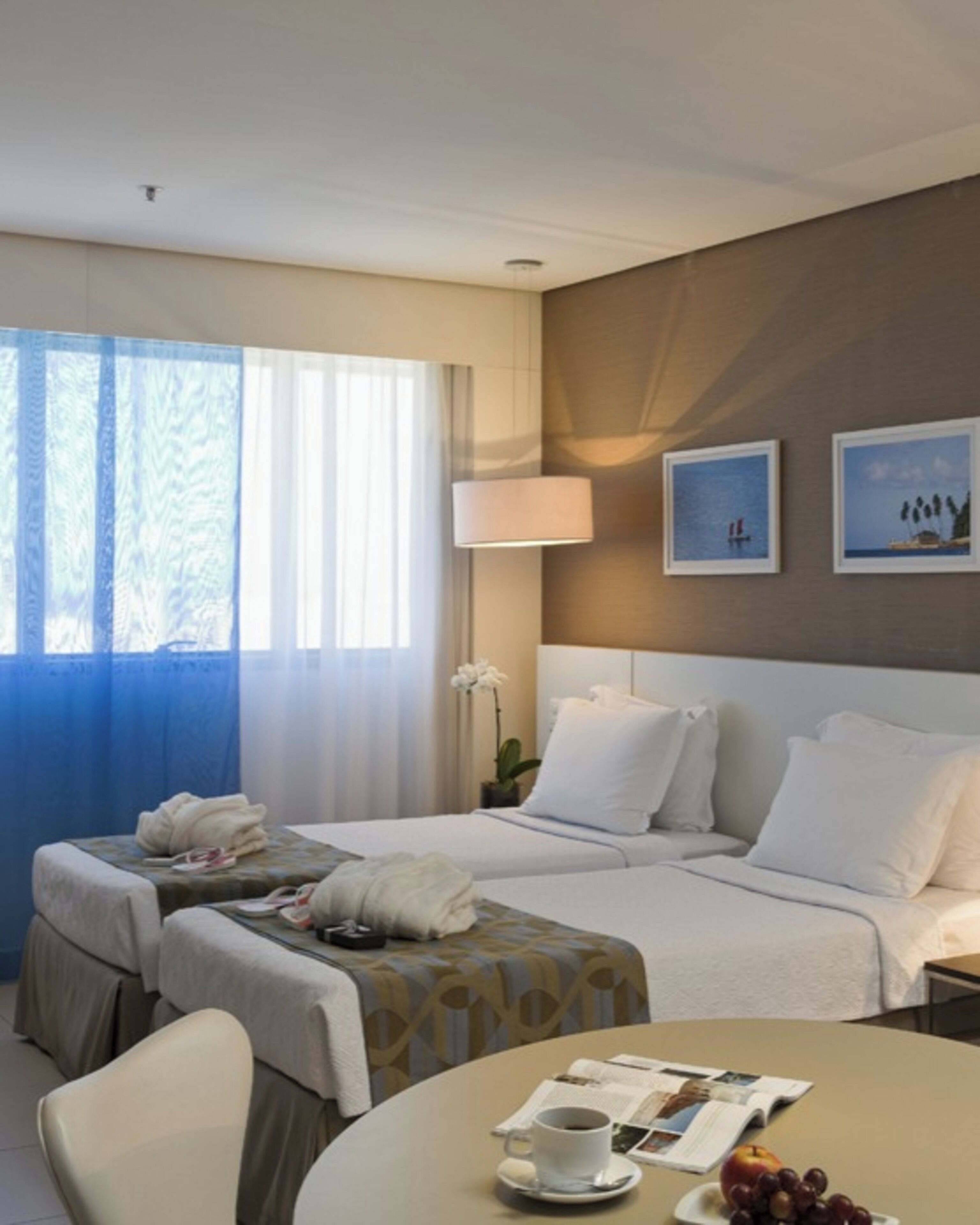 Quarto de hotel com duas camas, uma mesa redonda com uma xícara e frutas, e uma janela com cortinas.
