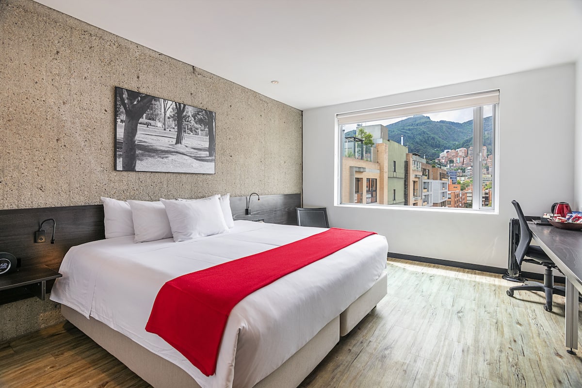 Quarto de hotel moderno com uma cama grande, vista para o horizonte urbano e manta vermelha.
