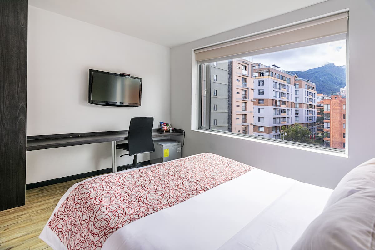 Quarto de hotel com cama, escrivaninha, cadeira, televisão e janela com vista para a cidade.