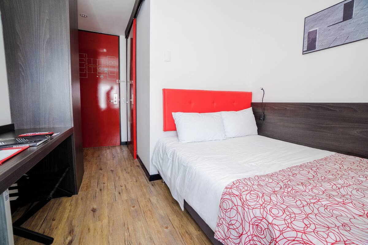 Quarto de hotel com chão de madeira, porta vermelha, cama com colcha branca e vermelha estampada, e escrivaninha com objetos vermelhos e pretos.