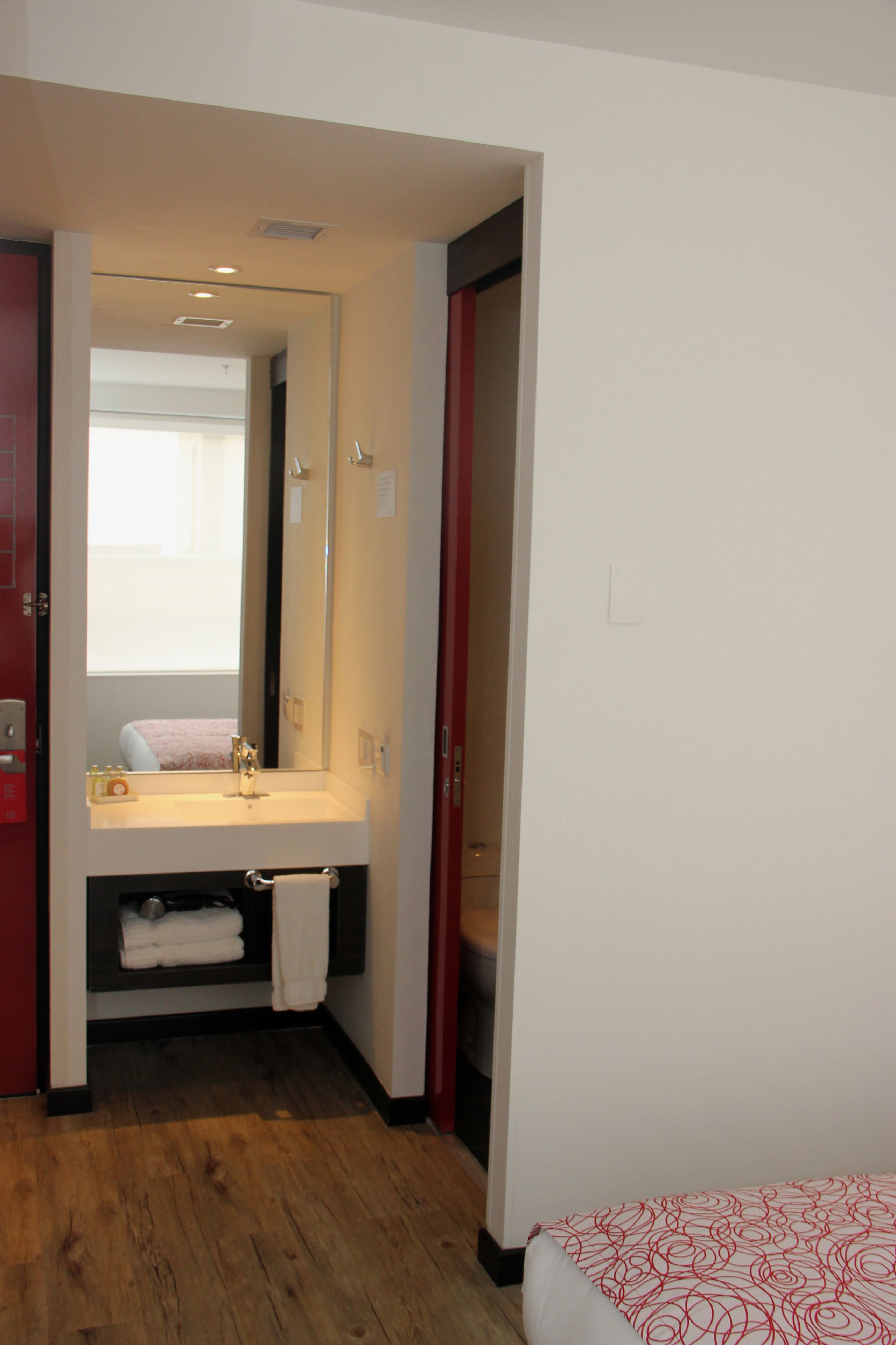Quarto de hotel com piso de madeira, cama branca com colcha estampada em vermelho e branco, piaça branca com espelho acima e porta vermelha.