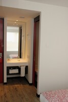 Quarto de hotel com piso de madeira, cama branca com colcha estampada em vermelho e branco, piaça branca com espelho acima e porta vermelha.