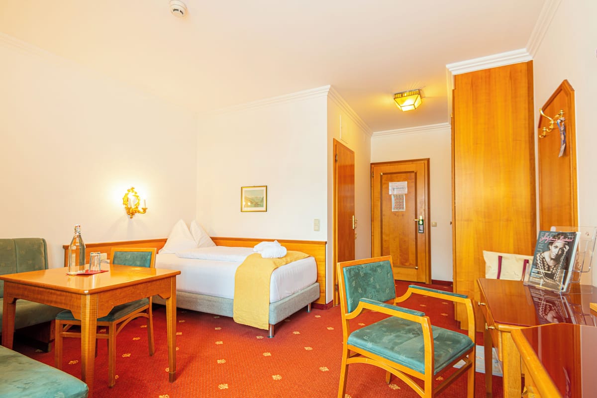Quarto de hotel com uma cama, uma escrivaninha com cadeira, uma mesa com cadeira e uma porta de madeira.