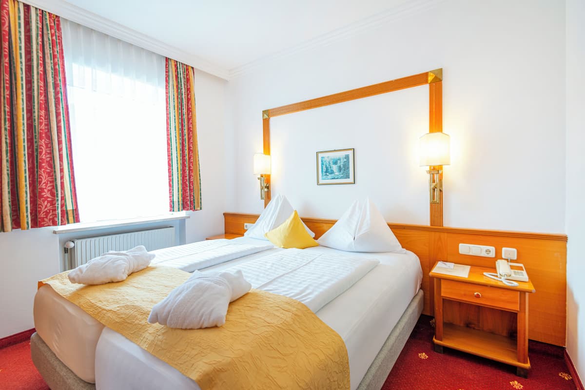 Quarto de hotel com uma cama grande, duas mesas de cabeceira, uma mesinha de cabeceira com abajur e uma janela com cortinas.