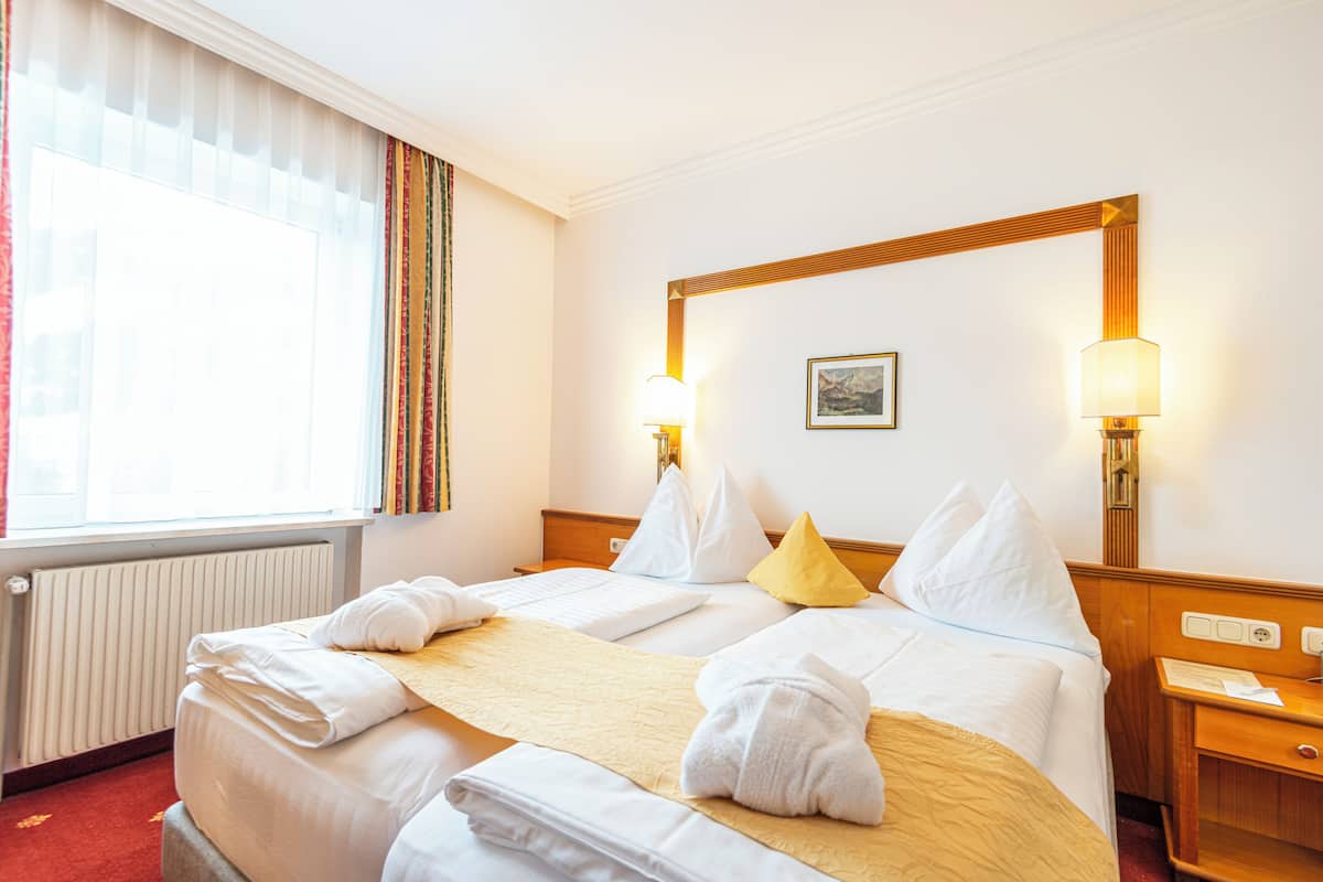 Quarto de hotel com duas camas, cabeceira de madeira, janela com cortinas e duas luminárias de cabeceira.