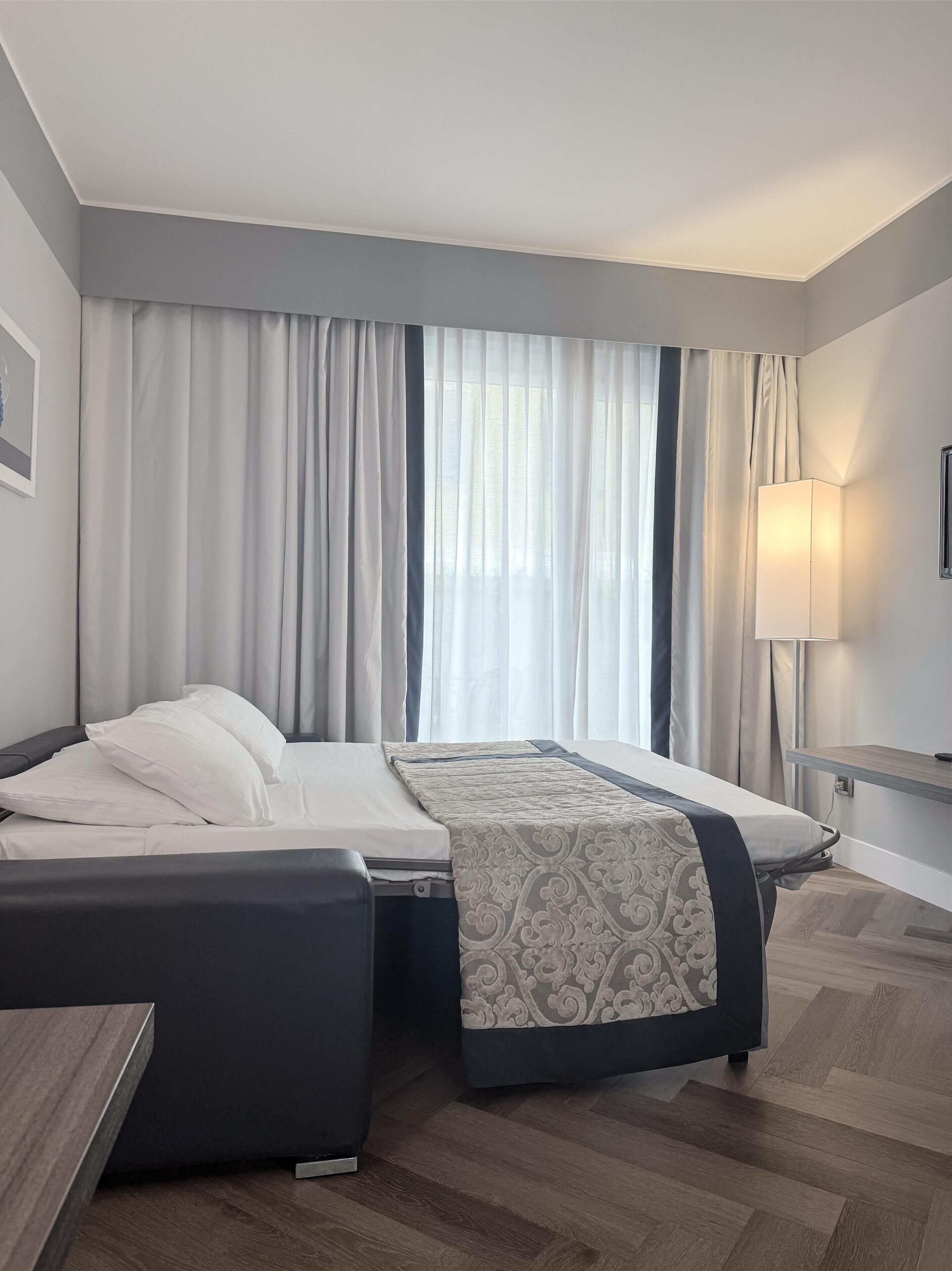 Quarto de hotel com duas camas, uma escrivaninha, uma luminária e uma janela grande com cortinas.