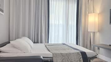 Quarto de hotel com duas camas, uma escrivaninha, uma luminária e uma janela grande com cortinas.