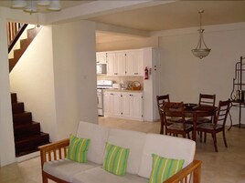 Uma sala de estar com sofá, cadeiras, uma mesa de jantar e uma cozinha ao fundo.