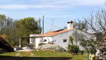 Uma casa rústica com telhado de telhas, chaminé de pedra e um pequeno jardim com um caminho que leva à entrada.