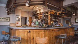 Uma área de bar com balcão de madeira, vários banquinhos e uma variedade de garrafas e copos.