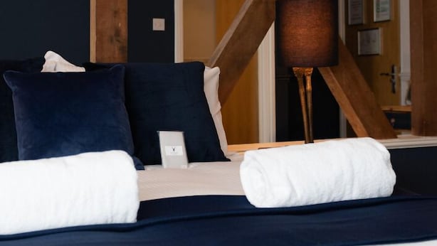 Cama com roupa de cama azul escura e almofadas brancas, estrutura de teto de madeira e um abajur no chão.