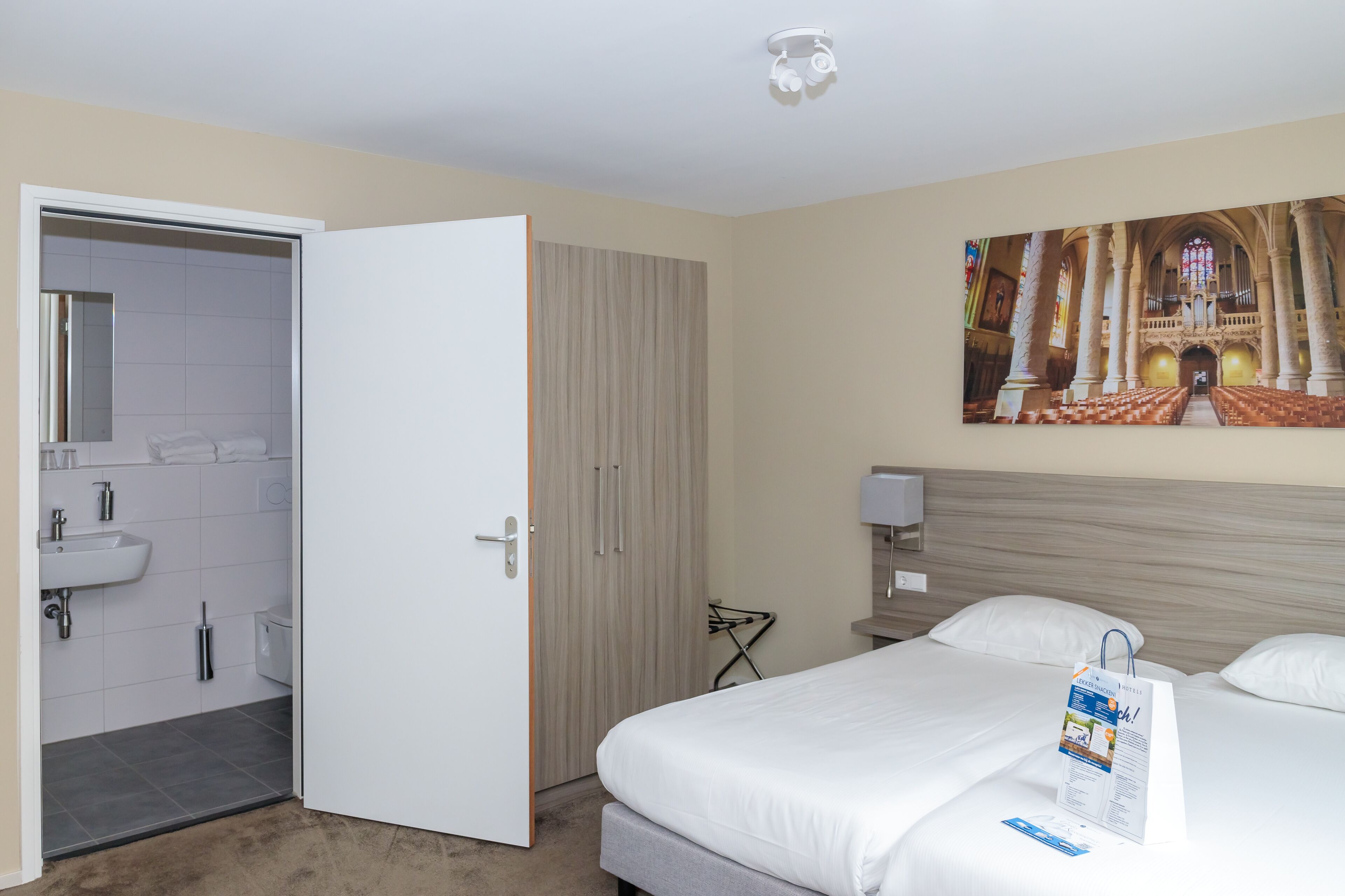 Quarto de hotel com uma cama, uma porta branca que dá para um banheiro, um quadro de uma catedral e um folheto sobre a cama.