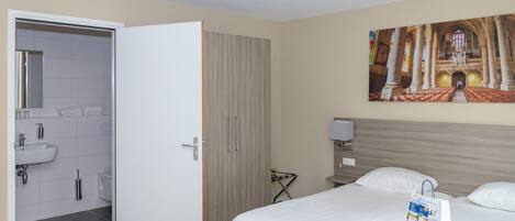 Quarto de hotel com uma cama, uma porta branca que dá para um banheiro, um quadro de uma catedral e um folheto sobre a cama.